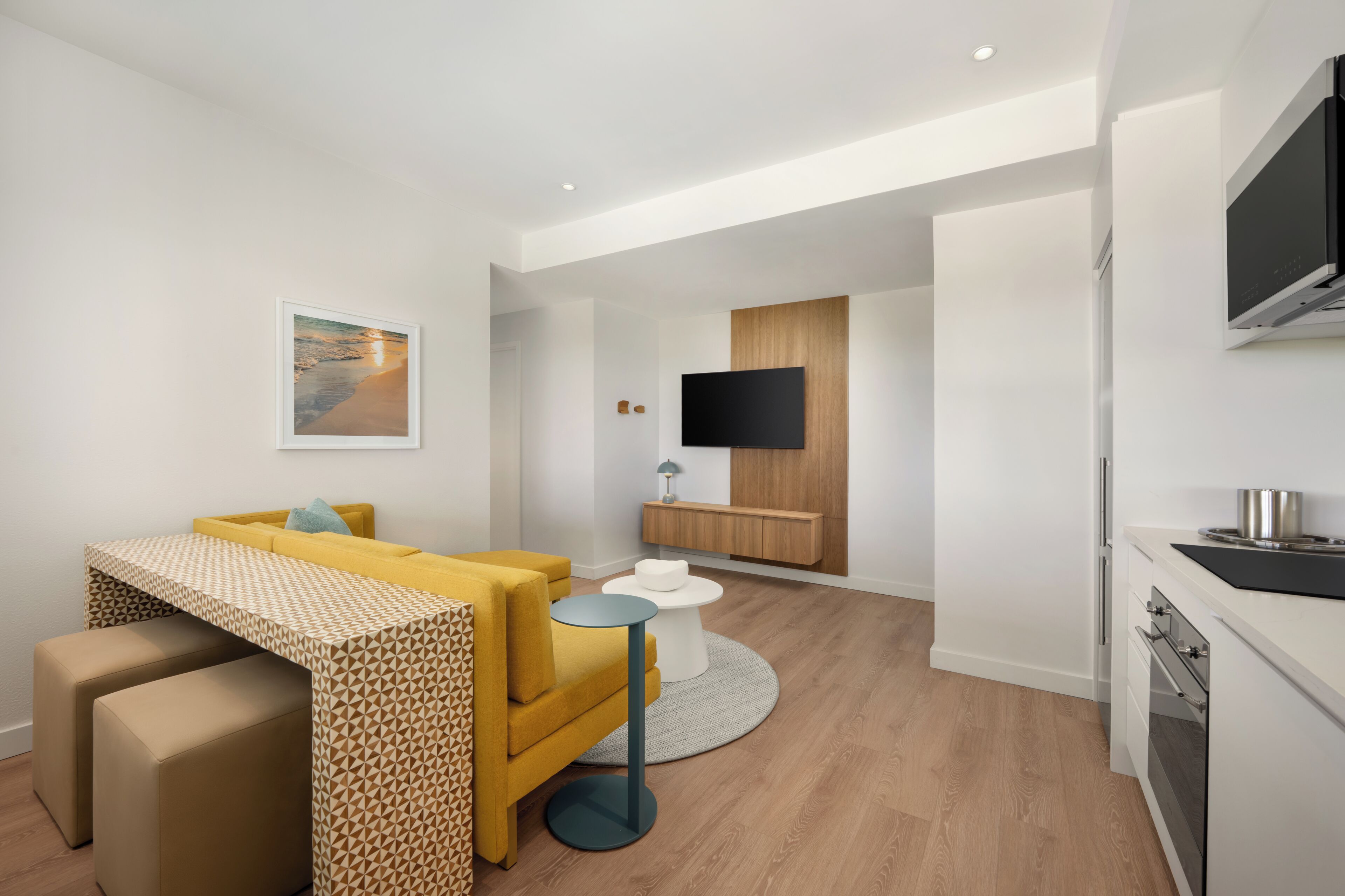 Uma sala de estar moderna com sofá, mesa de centro e um banquinho, uma mesinha lateral redonda, uma TV de tela plana fixada em um painel de madeira e uma área de cozinha com fogão e forno.