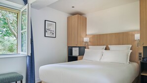 Quarto de hotel moderno com uma cama grande, uma mesa de cabeceira com luminária, um pequeno banco e uma janela com cortinas.