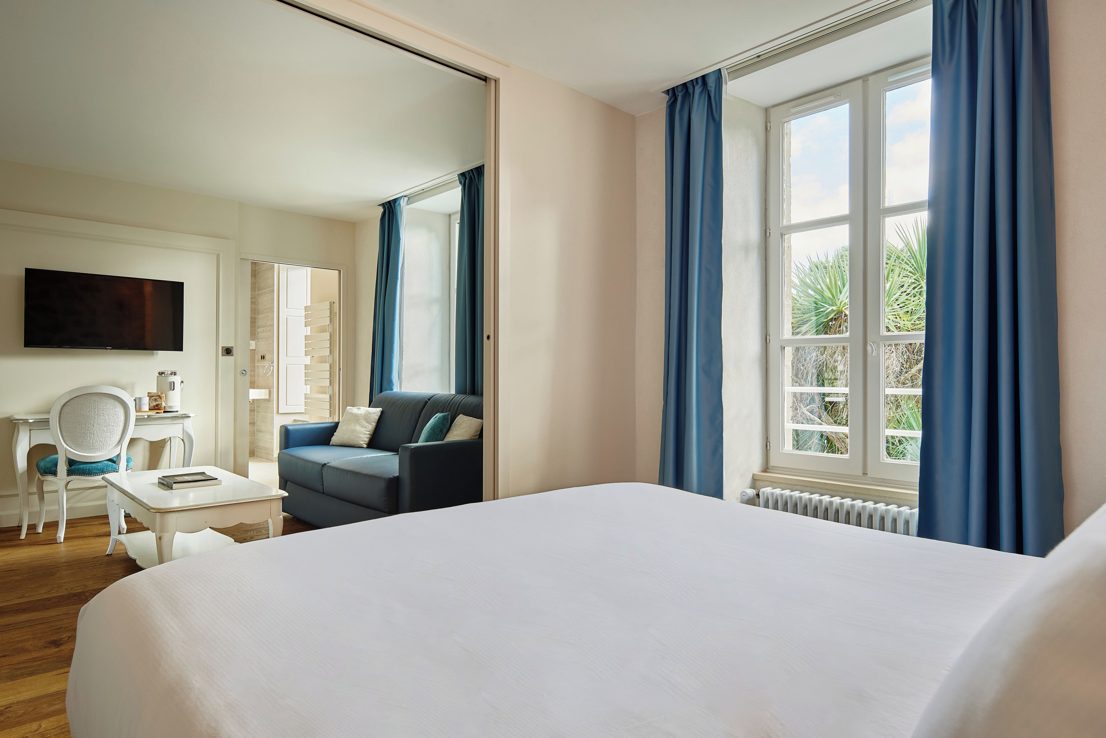 Quarto de hotel com uma cama grande, uma escrivaninha com cadeira, um sofá, uma televisão e uma janela com cortinas.