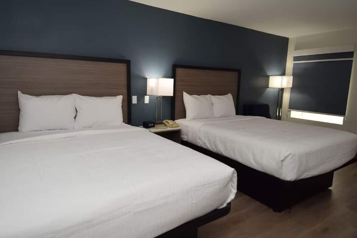 Quarto de hotel com duas camas, cada uma com cabeceira de madeira, roupa de cama branca e abajures.