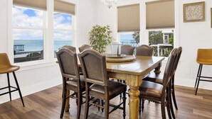 Uma sala de jantar com uma mesa grande, seis cadeiras, um lustre e janelas amplas com vista para o oceano.