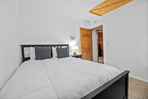 Quarto com cama, mesa de cabeceira, abajur e porta de madeira que dá para outro cômodo.