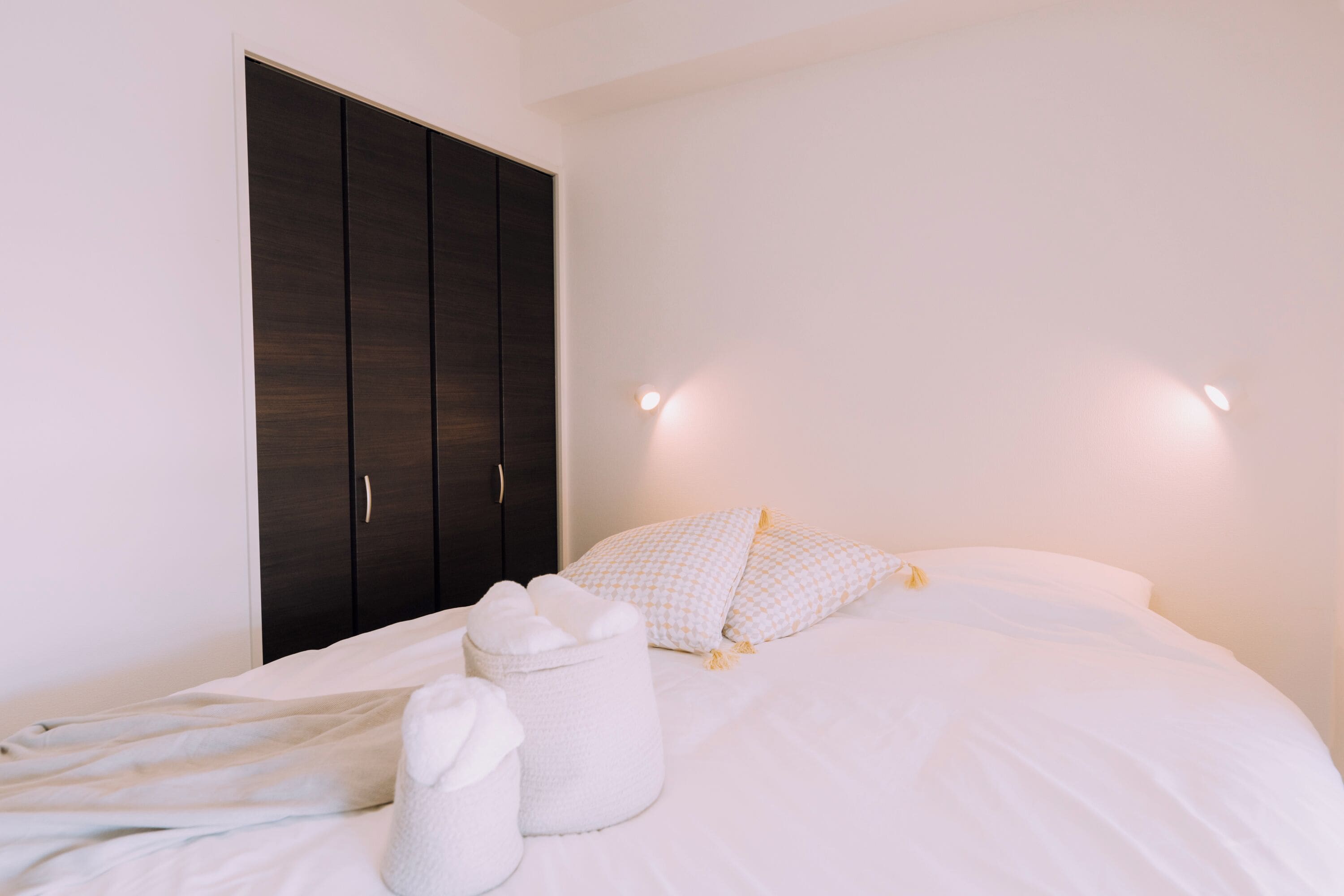 Cama bem arrumada com lençóis brancos, guarda-roupa escuro e uma pequena mesa de cabeceira com toalhas.