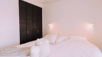 Cama bem arrumada com lençóis brancos, guarda-roupa escuro e uma pequena mesa de cabeceira com toalhas.