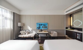 Quarto de hotel com duas camas, uma escrivaninha com luminária, uma TV exibindo cartazes de filmes e um espelho grande com iluminação.