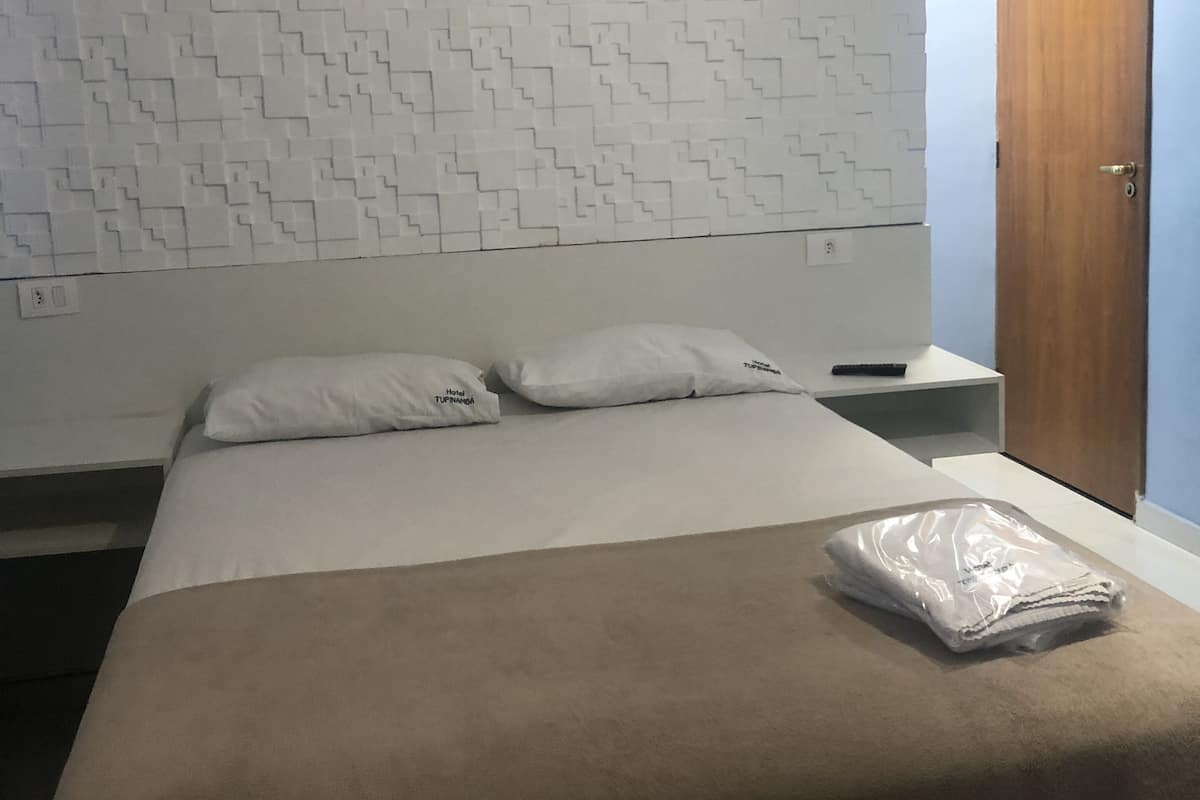 Quarto de hotel com parede estampada, cama com manta bege, duas almofadas, mesa de cabeceira com controle remoto e porta de madeira.