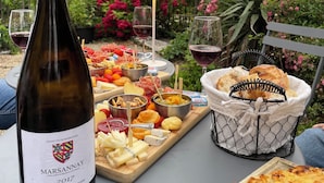 Uma garrafa de vinho com o rótulo "Marsannay 2017" sobre uma mesa, acompanhada de uma tábua de frios, queijos e cestos de pão.