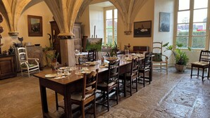 Uma sala de jantar com uma longa mesa de madeira preparada para seis pessoas, cercada por cadeiras de madeira. O espaço possui vigas de madeira aparente no teto, amplas janelas e diversas obras de arte nas paredes.