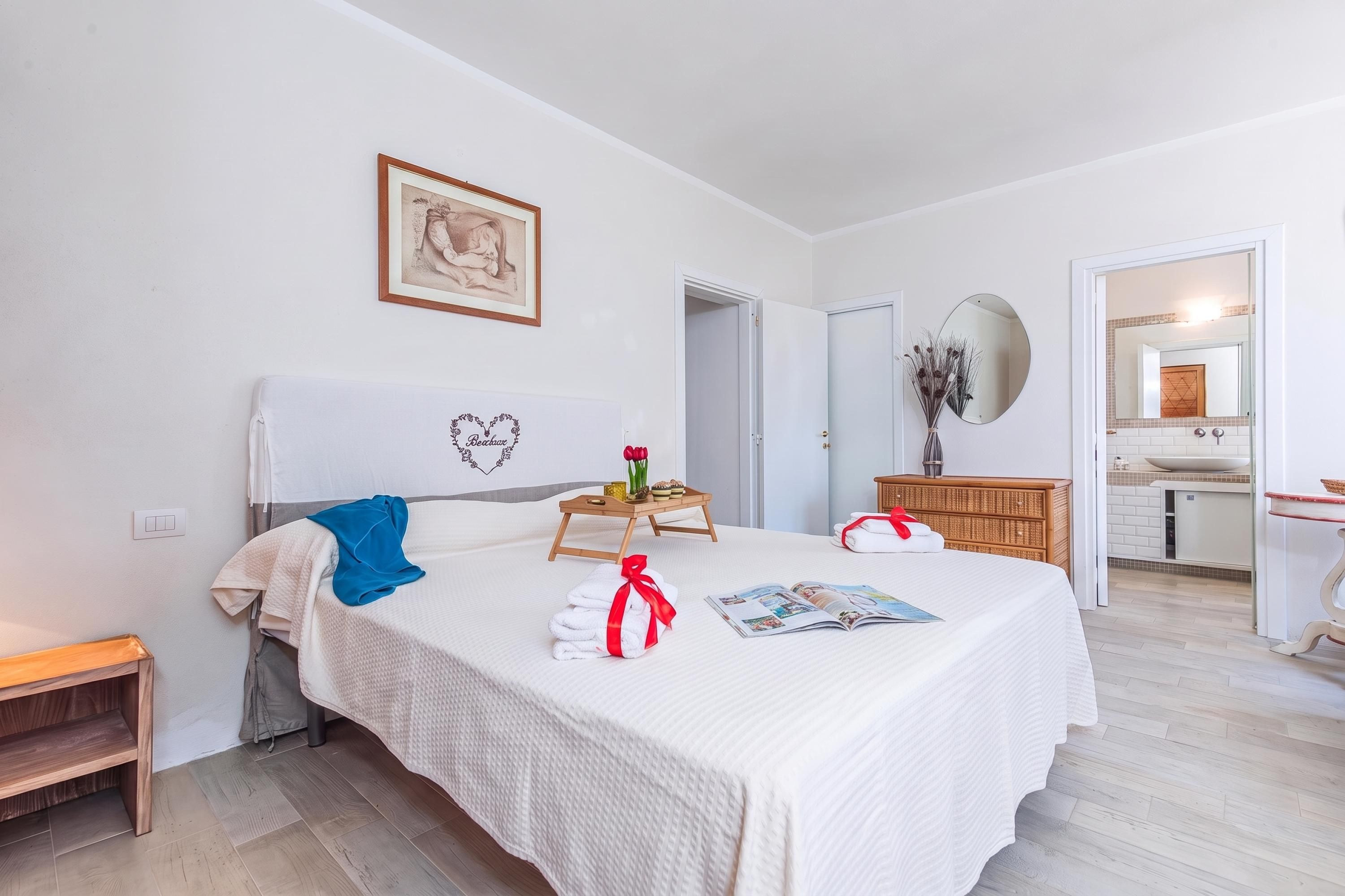 Quarto de hotel com cama, mesa de cabeceira, espelho, pequena mesa com um vaso de flores e porta que dá para um banheiro.