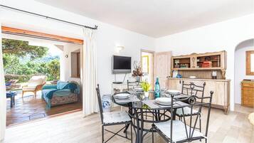 Uma sala de jantar com uma mesa posta para quatro pessoas, um sofá e uma televisão ao fundo.
