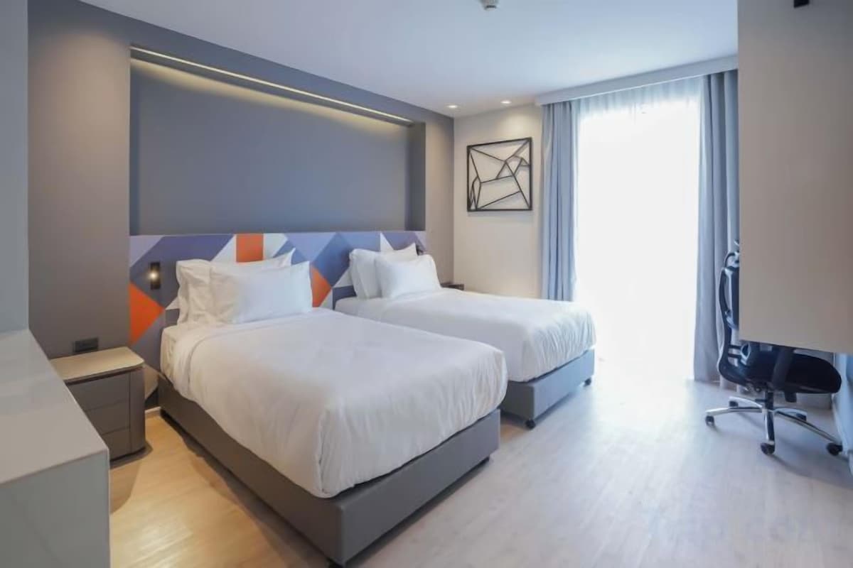 Quarto de hotel moderno com duas camas, uma escrivaninha e uma janela grande.