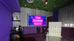 Quarto com letreiro de neon que diz 'THE SOIREE SUITE', área de bar com copos e garrafas, e parede verde com planta.