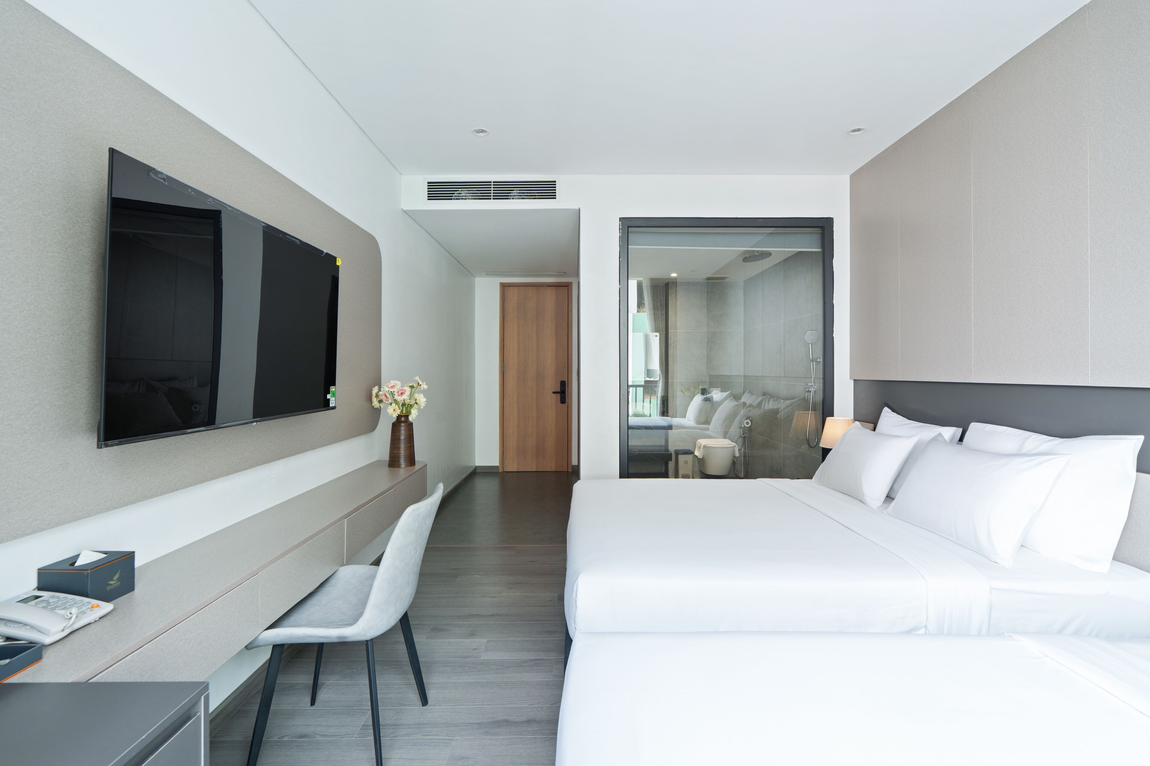Quarto de hotel moderno com uma cama grande, uma escrivaninha com cadeira, uma TV na parede e um banheiro visível através de uma porta de vidro.