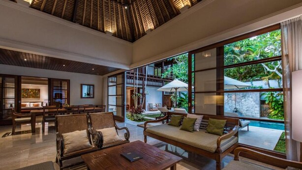 Espaço amplo e interno com teto alto, móveis de madeira e grandes janelas que dão para uma piscina e vegetação.