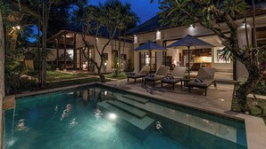 Área de piscina com espreguiçadeiras e guarda-sóis, cercada por plantas tropicais e um edifício moderno com grandes janelas.