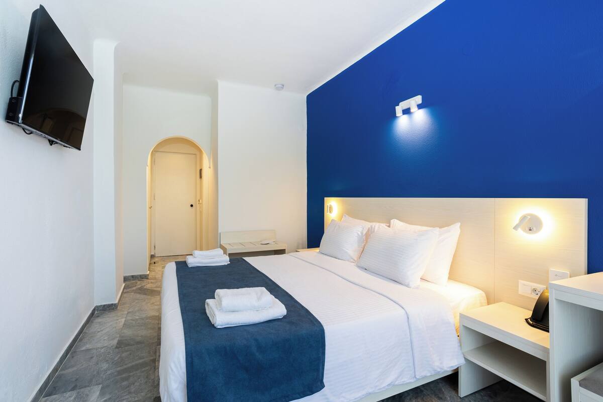 Quarto de hotel com cama, mesa de cabeceira, televisão na parede e porta para outro cômodo.