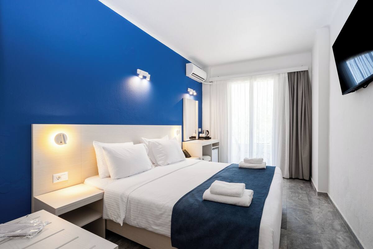 Quarto de hotel com uma cama grande, uma mesinha de cabeceira, uma escrivaninha, um espelho, uma televisão e uma janela com cortinas.