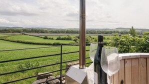Um deck de madeira com uma grelha de metal, uma garrafa de vinho e um copo, com vista para uma paisagem verde com campos e colinas ao longe.