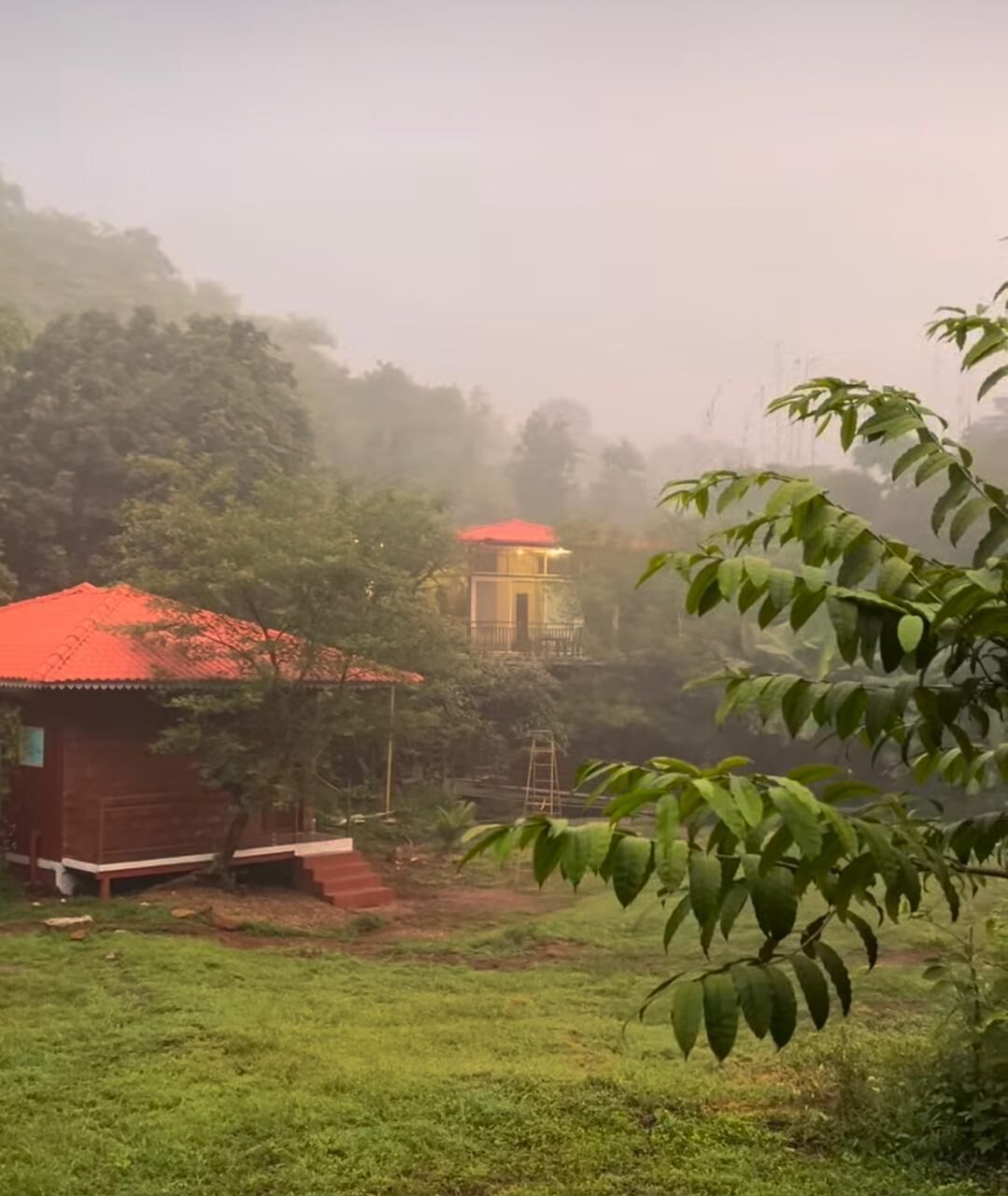 Uma casa com telhado vermelho e uma pequena construção também com telhado vermelho, numa área arborizada e com névoa.