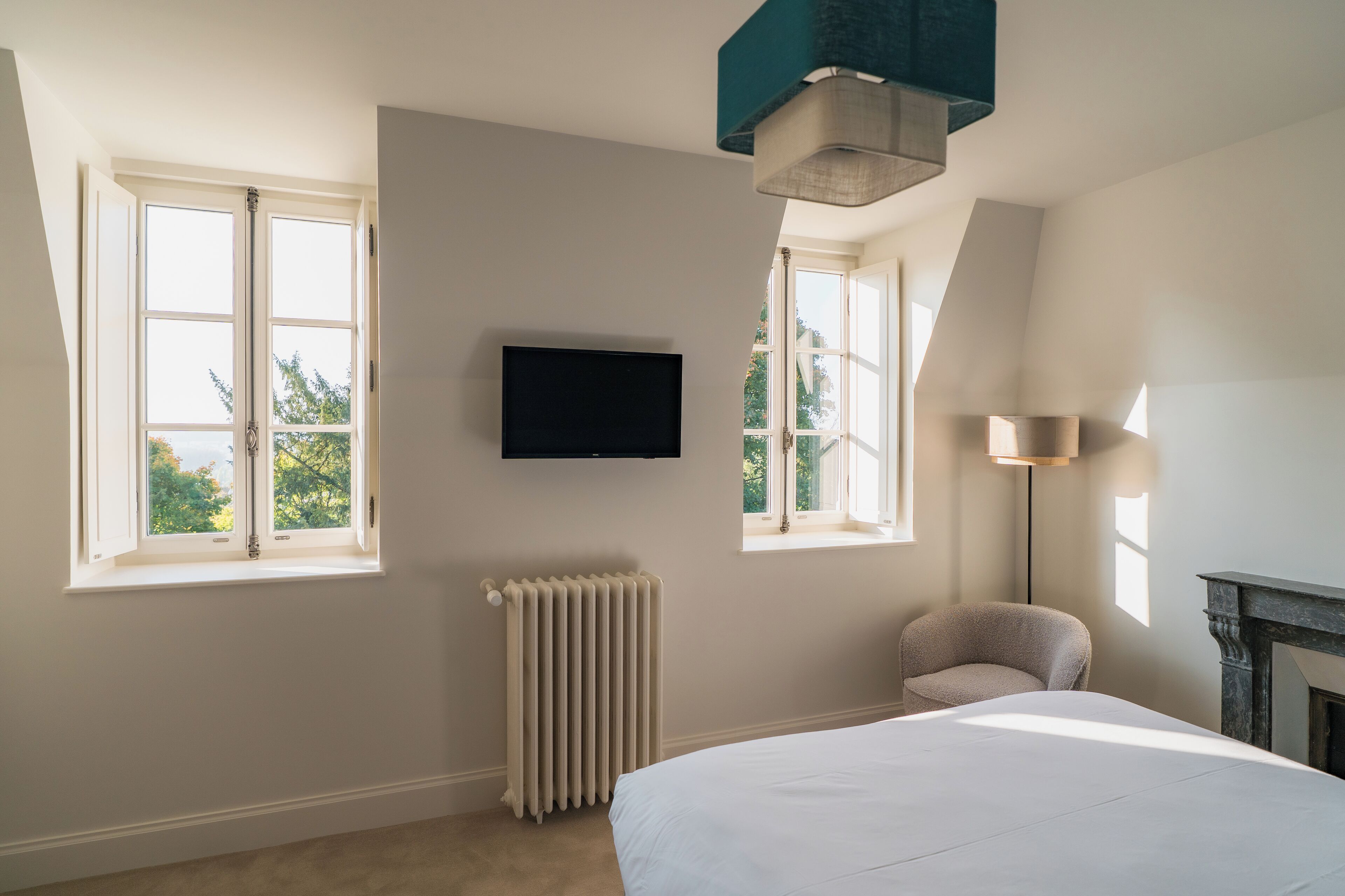 Quarto com cama, televisão presa à parede, radiador e duas janelas.