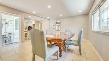 Uma área de refeições com mesa de madeira e quatro cadeiras estofadas, cozinha ao fundo e uma janela grande com persianas.