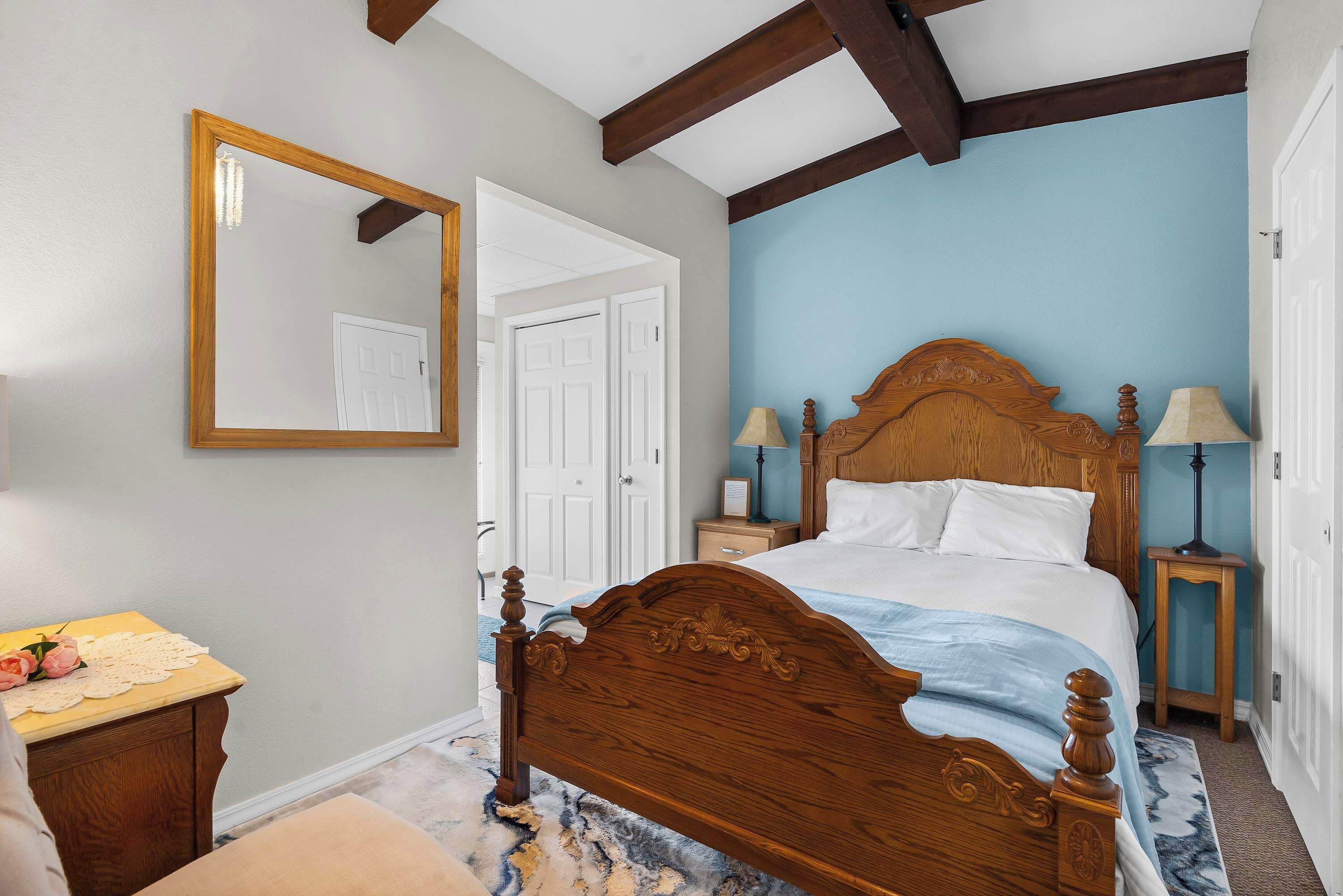Quarto com cama de madeira, mesa de cabeceira com luminárias, espelho e porta.