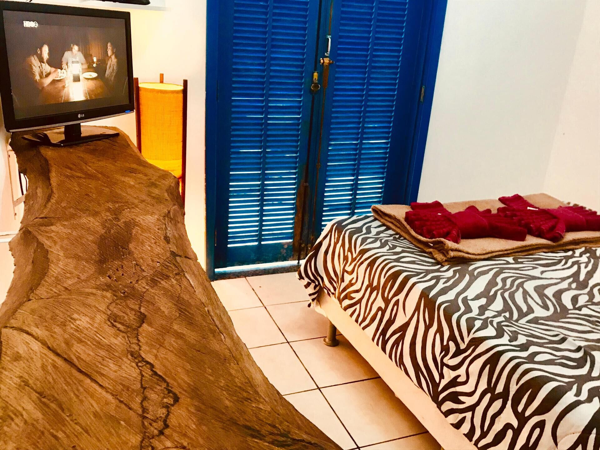 Quarto com televisão em suporte de madeira, cama com cobertura estampada de zebra e porta azul com persianas.