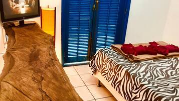Quarto com televisão em suporte de madeira, cama com cobertura estampada de zebra e porta azul com persianas.