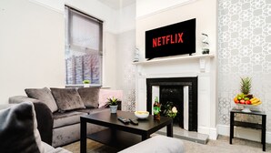 Uma sala de estar moderna com lareira, uma televisão exibindo 'NETFLIX', uma mesa de centro com controles remotos e uma cesta de frutas em uma mesinha lateral.