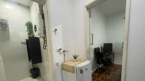 Cozinha compacta com paredes revestidas de azulejos brancos, uma geladeira, uma pia pequena e uma planta em vaso.