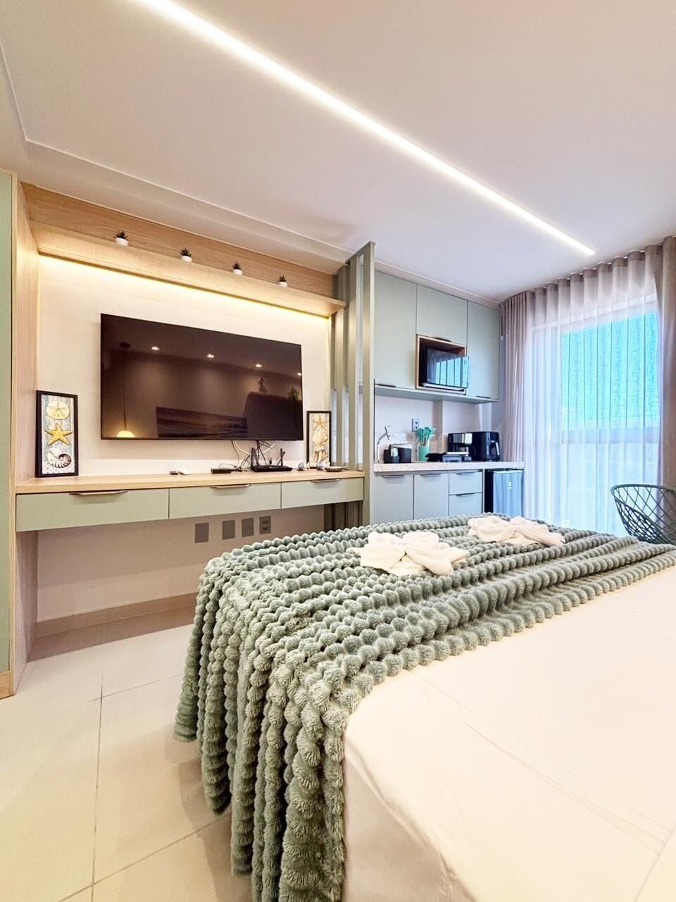 Quarto de hotel moderno com cama grande, televisão de tela plana, cozinha compacta e uma janela ampla com cortinas.