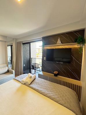 Quarto de hotel com uma cama grande, televisão de tela plana fixada em parede de mármore, varanda com uma cadeira e vista para o exterior.