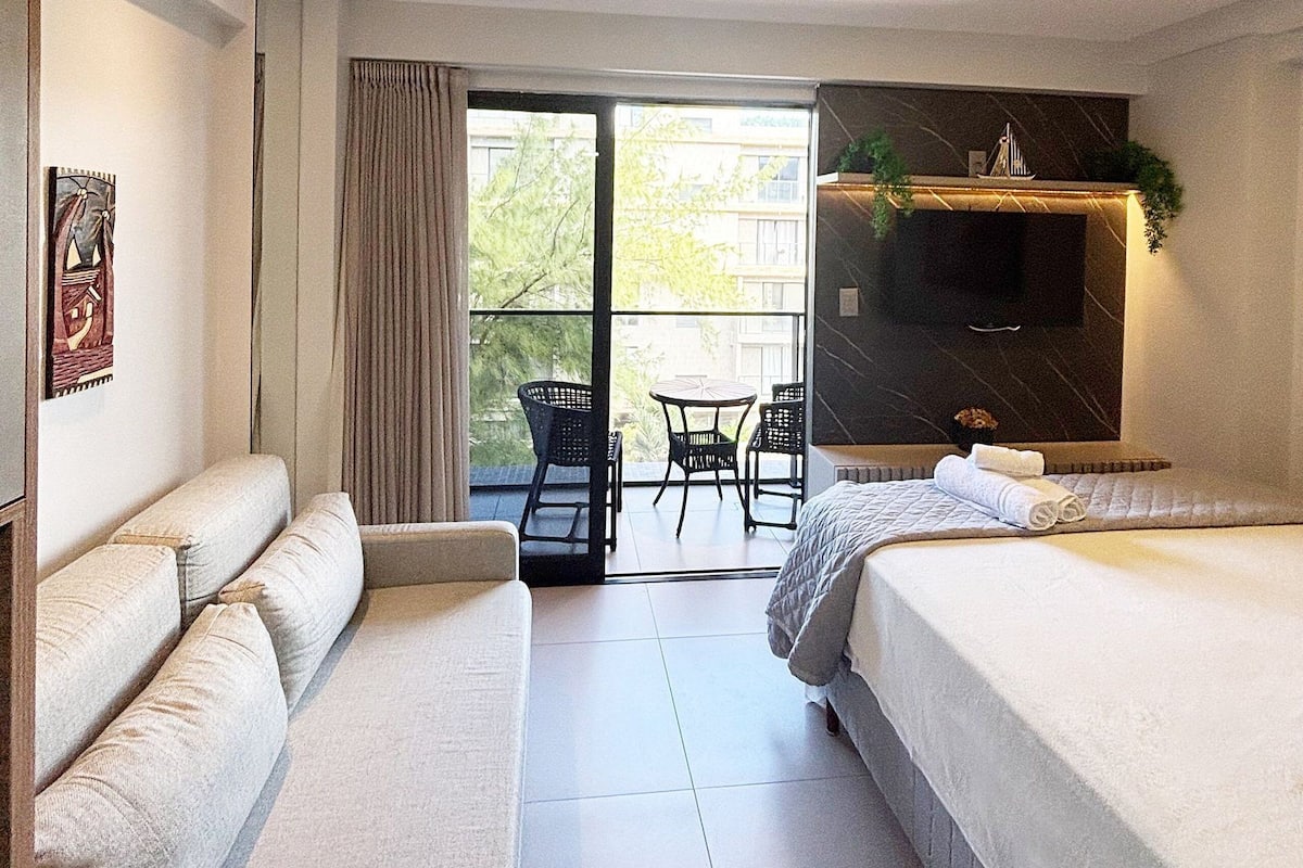 Quarto de hotel com sofá, cama, televisão, varanda com cadeiras e vista para o verde.