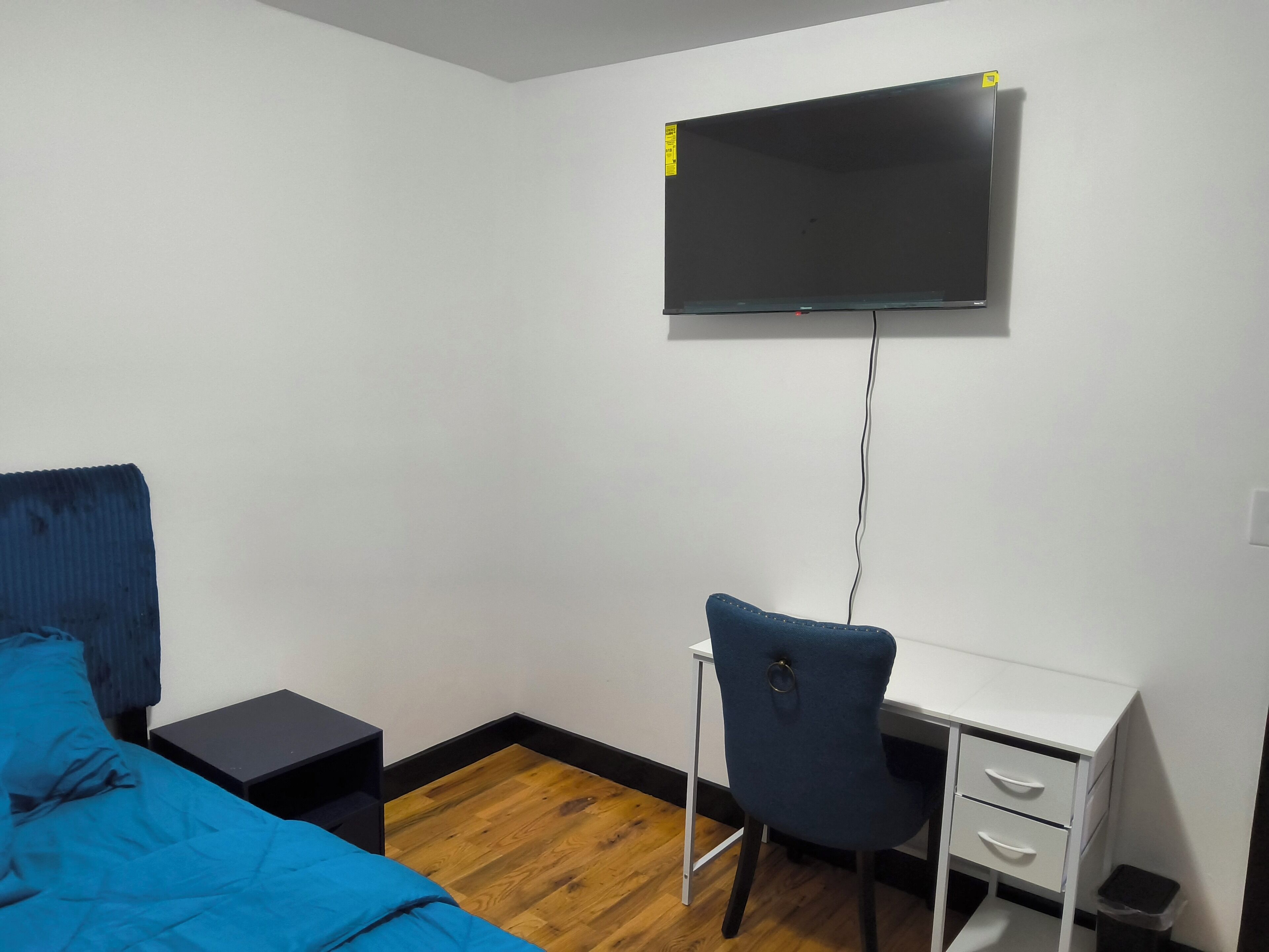 Quarto com cama, escrivaninha, cadeira e televisão fixada na parede.