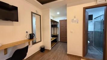 Quarto de hotel com chão de madeira, uma cama, uma mesa pequena com chaleira, um espelho e uma TV fixada na parede.