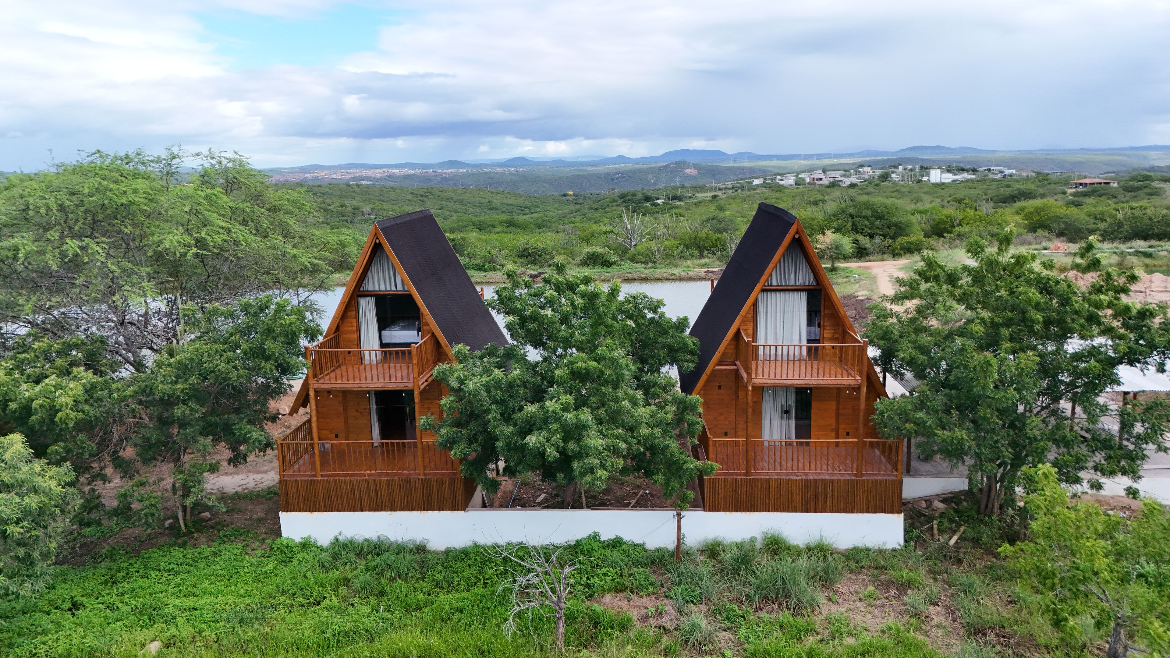 Duas cabanas de madeira com varandas, cercadas por vegetação e um lago, com vista para colinas ao longe.