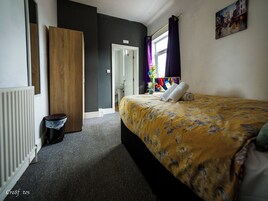 Quarto de hotel com cama, radiador, guarda-roupa de madeira, lixeira e janela com cortinas.