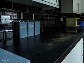 Cozinha com bancada preta, três recipientes identificados para açúcar, café e chá, micro-ondas e uma tomada.