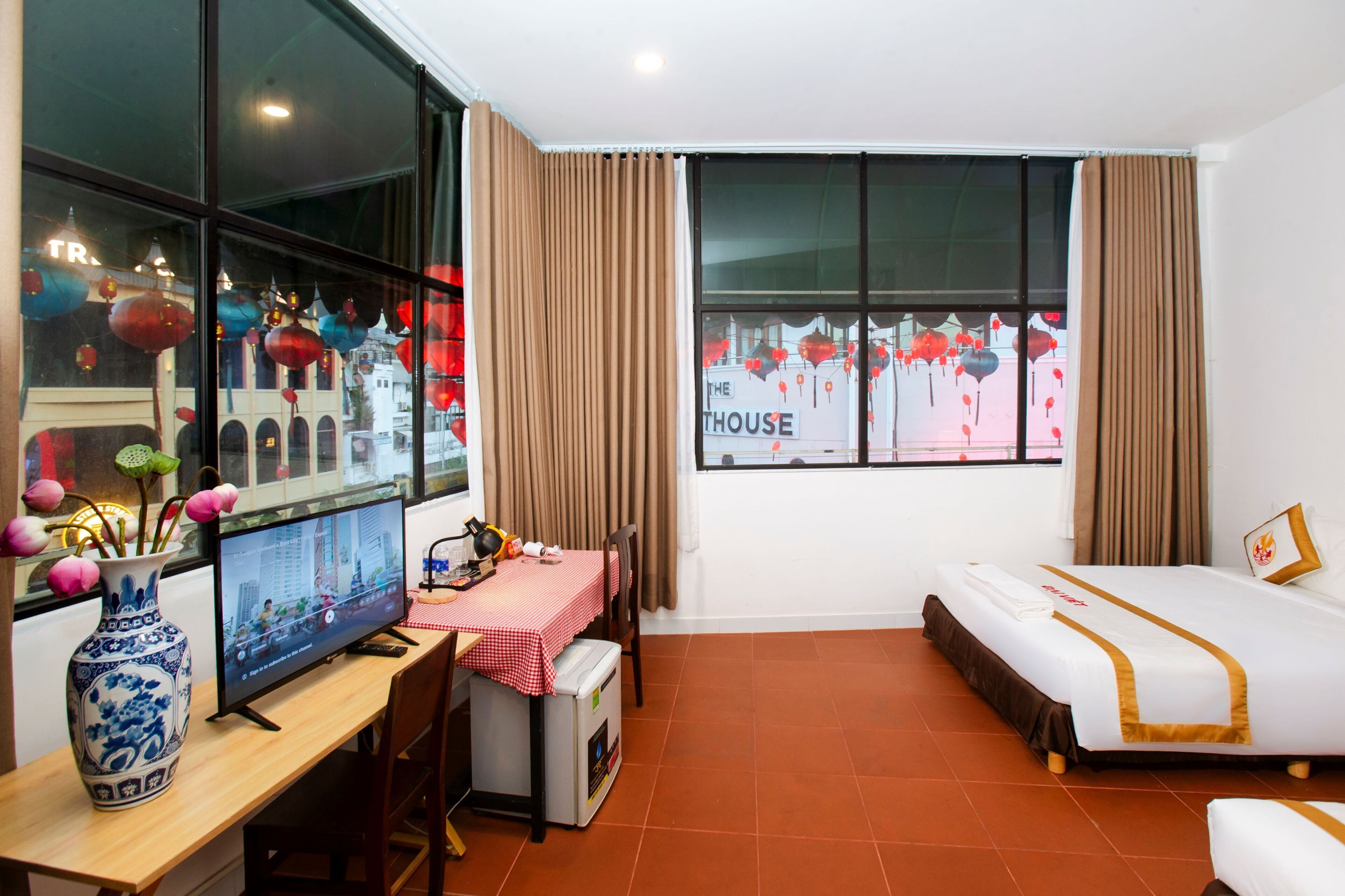 Quarto de hotel com cama, escrivaninha com computador, mesinha, televisão e janela com cortinas.