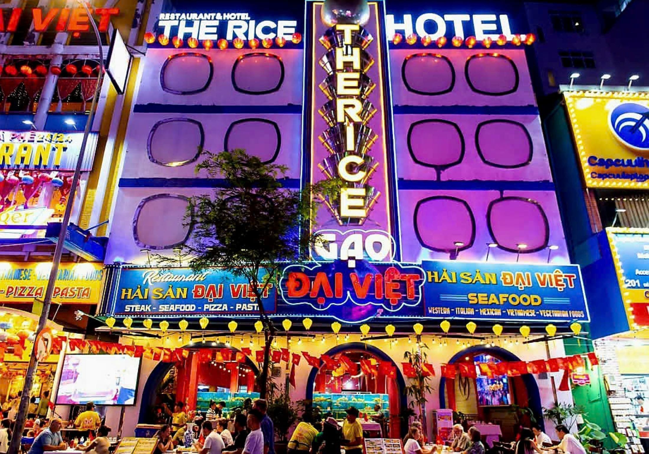 A brightly lit building with multiple signs, including "The Rice Hotel" and "Hai San Dai Viet Seafood."