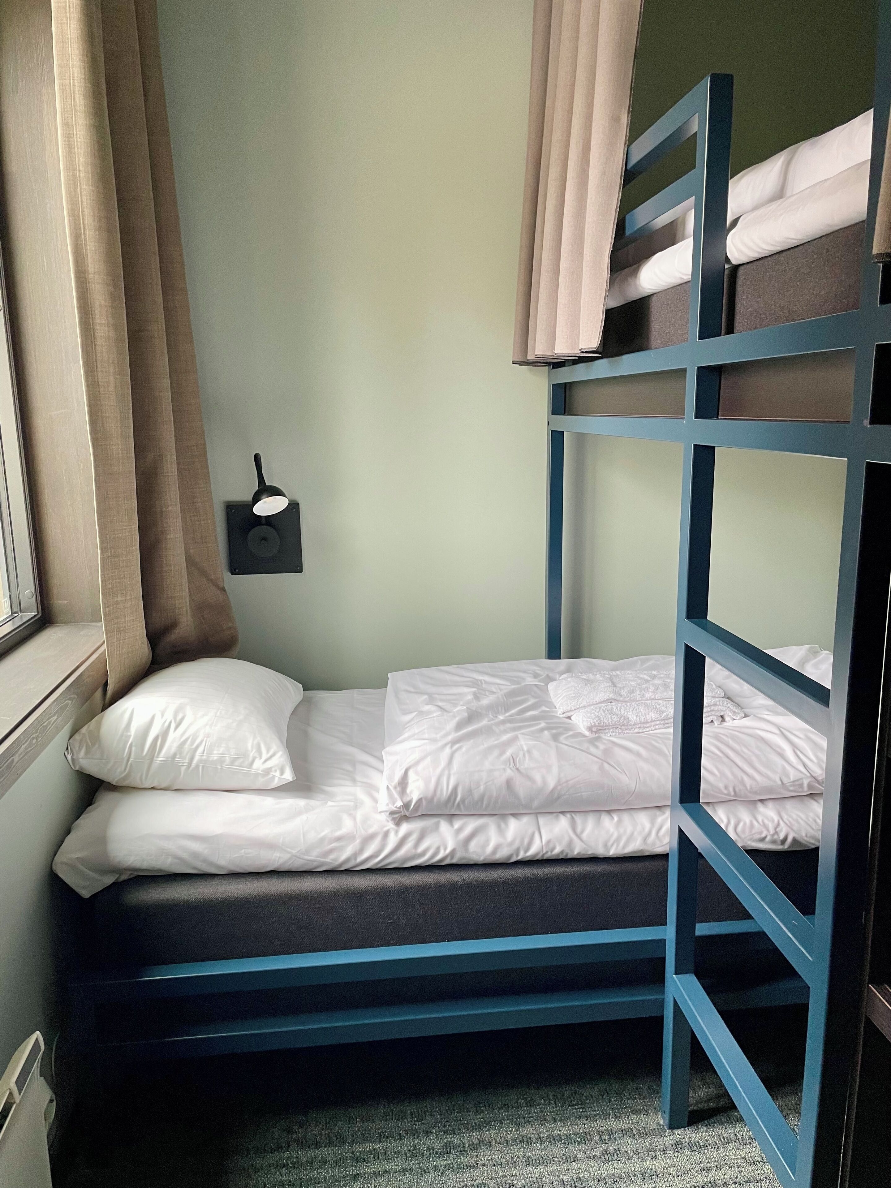 Uma cama beliche com roupa de cama branca, uma janela com cortinas bege e um interruptor de luz fixado na parede.