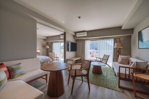 Quarto de hotel com sofá, duas mesas de madeira, televisão e uma janela grande com cortinas.