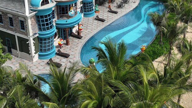 Resort com uma grande piscina cercada por palmeiras e uma área externa coberta com espreguiçadeiras.