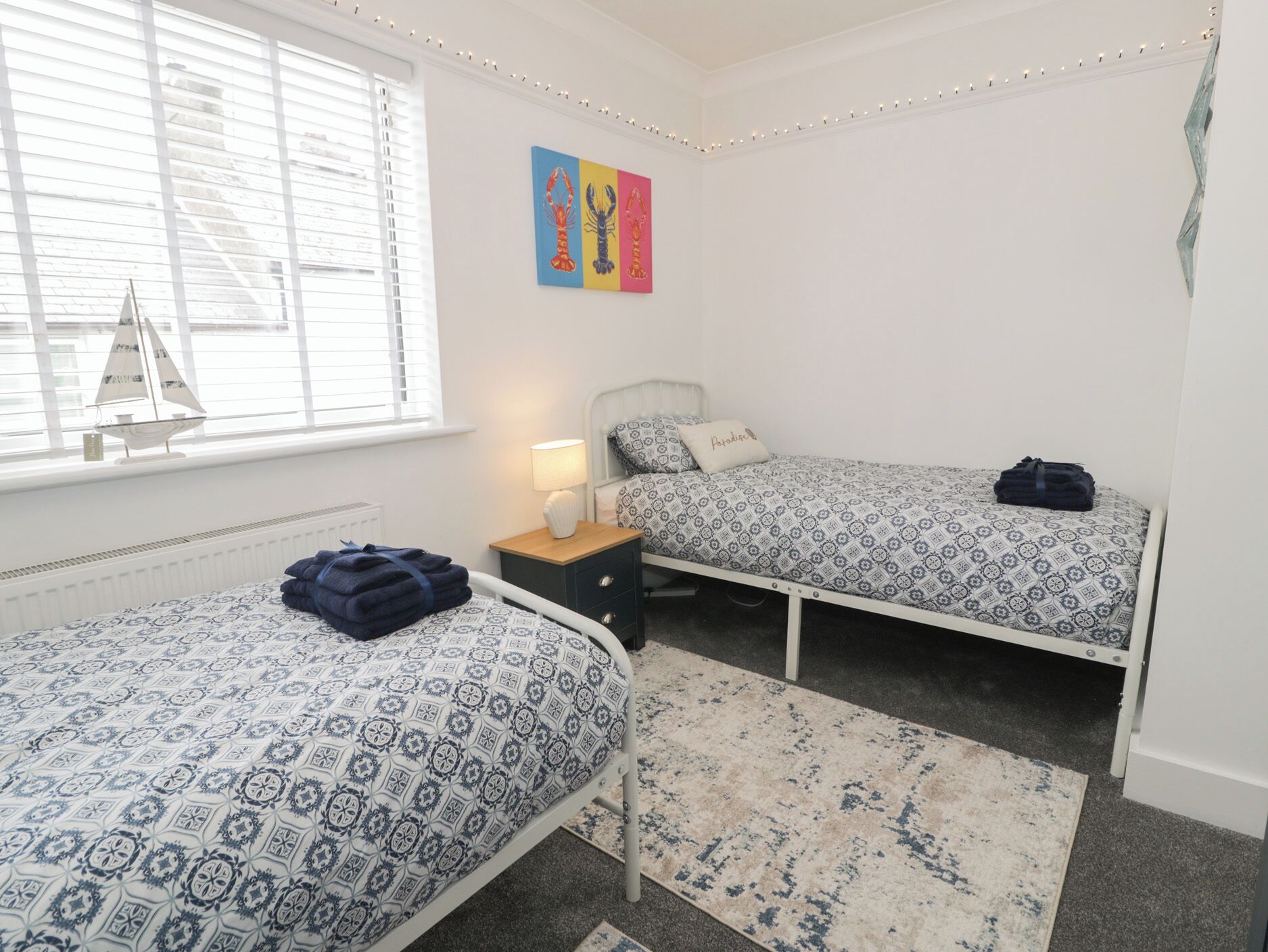 Quarto com duas camas, mesa de cabeceira, luminária, decoração náutica de um veleiro e uma obra de arte colorida na parede.