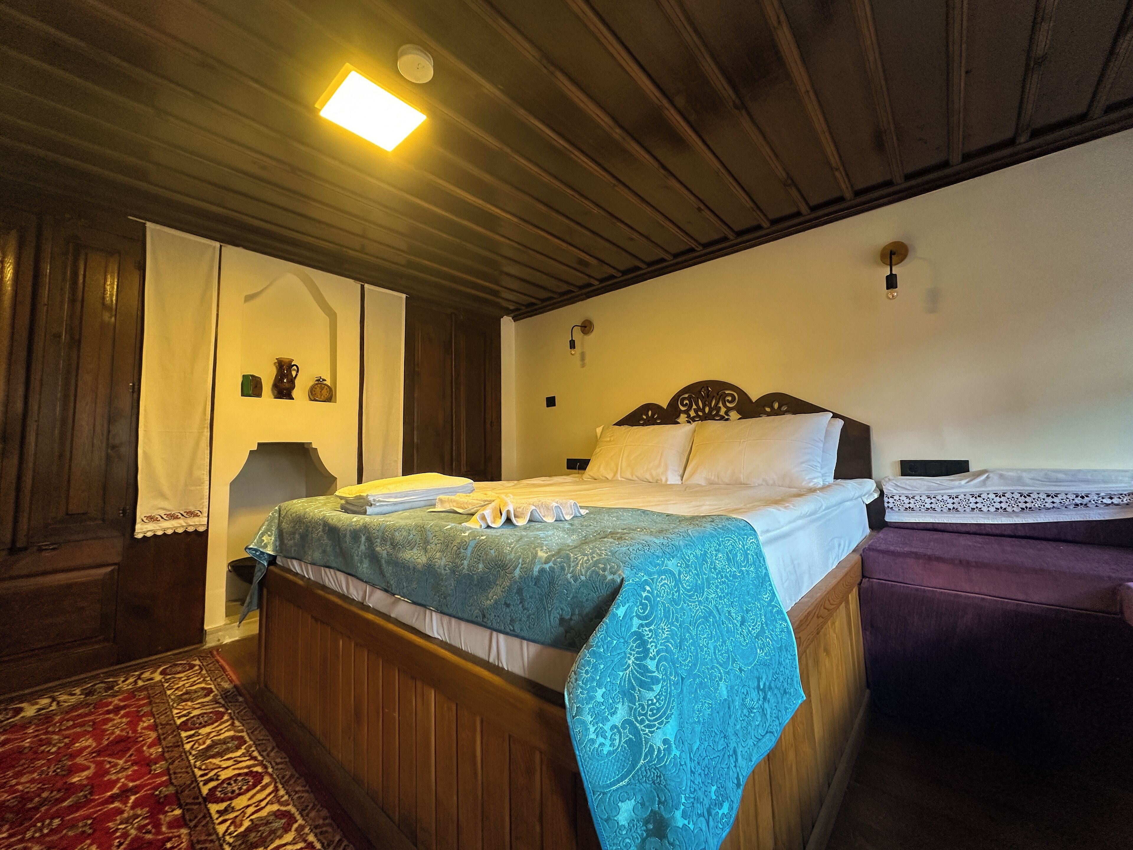 Quarto com cama de madeira, mesa de cabeceira, luminária na parede e tapete estampado.