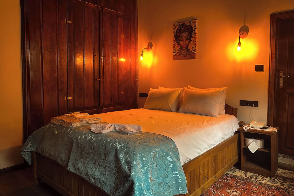 Cama bem arrumada com colcha azul estampada, guarda-roupa de madeira e duas luminárias na parede.