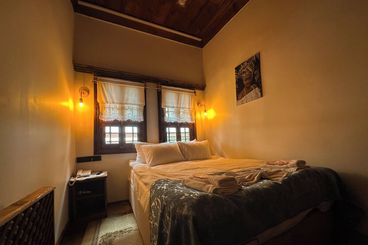 Quarto com cama, mesa de cabeceira, abajur e janela com cortinas.
