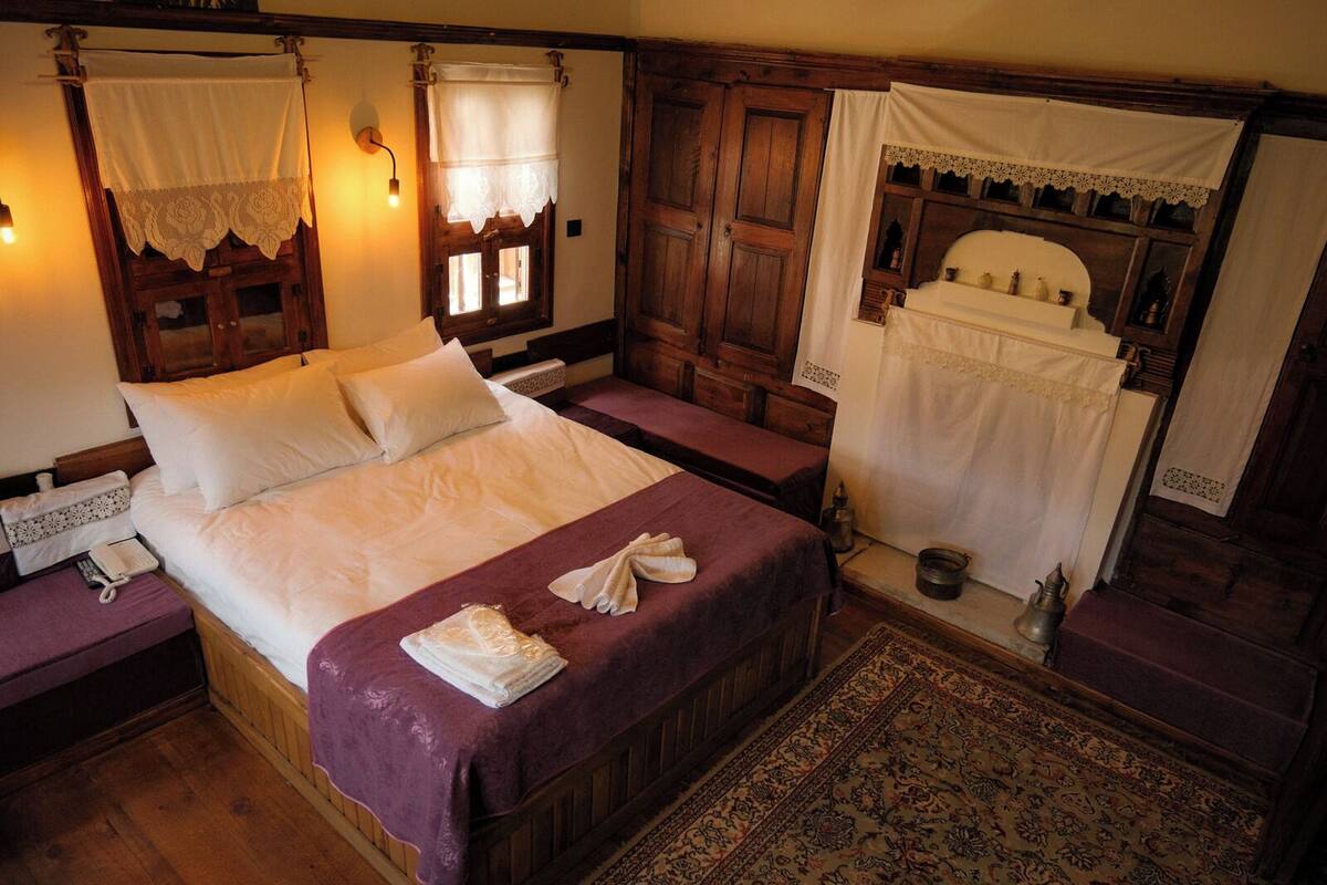 Quarto tradicional com cama grande, guarda-roupa de madeira e pequena mesa com abajur.