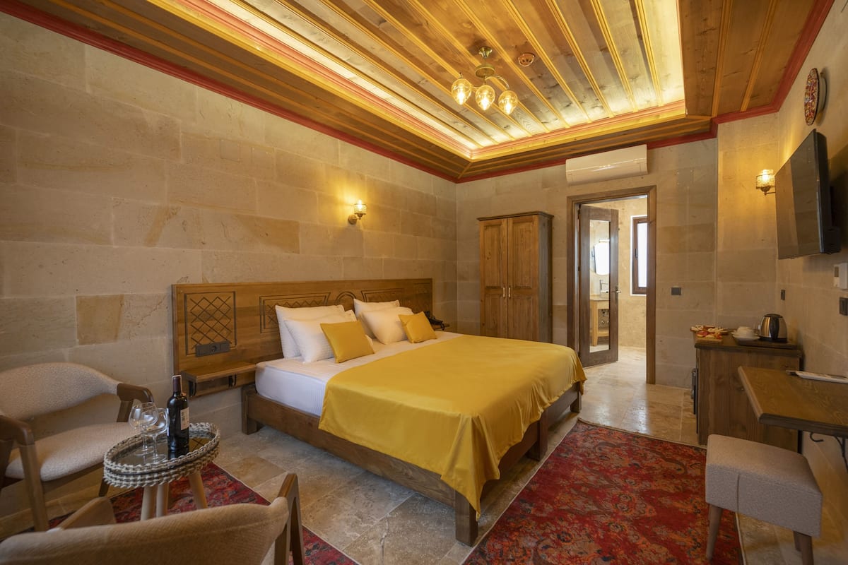 Quarto de hotel com uma cama grande, cabeceira de madeira, uma mesinha com garrafa e copo, e uma televisão na parede.