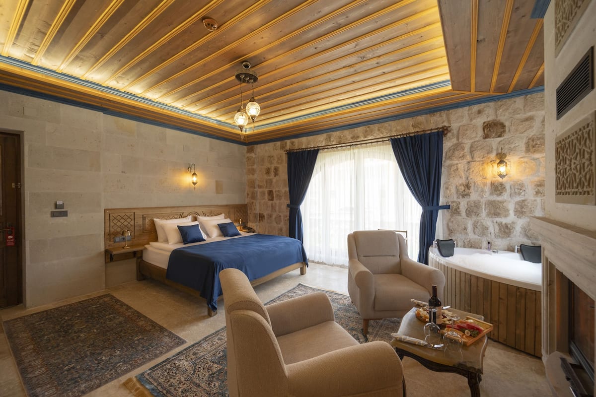 Quarto com teto de madeira, paredes de pedra, cama com roupa de cama azul, área de estar com cadeiras e uma pequena mesa com garrafa e petiscos.
